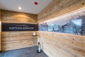 Uma área de recepção com painéis de madeira, uma grande moldura com uma paisagem nevada e uma placa com a inscrição 'ANTHOLZERHOF'.