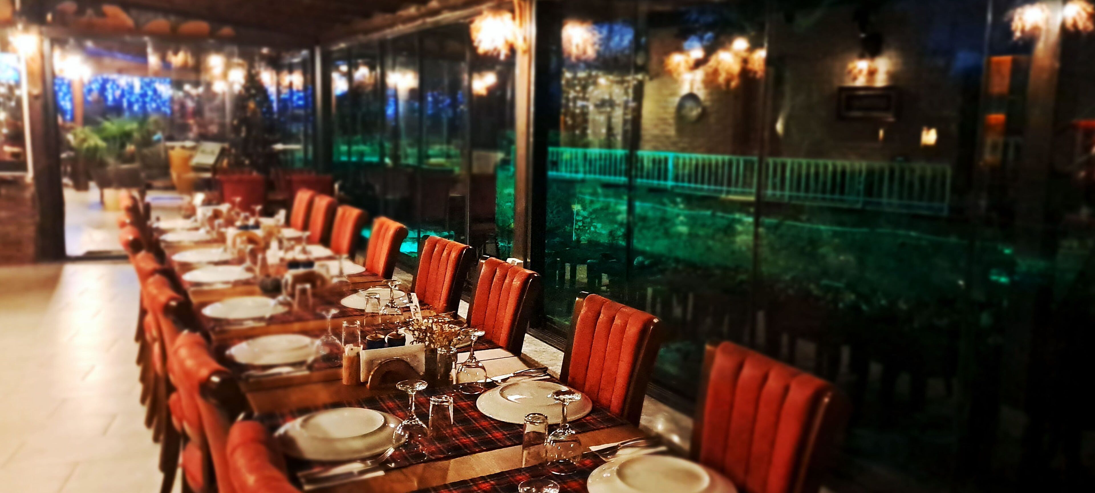Restaurante com uma mesa comprida posta para refeições, cadeiras estofadas vermelhas e vista para área externa iluminada em verde através de grandes janelas.