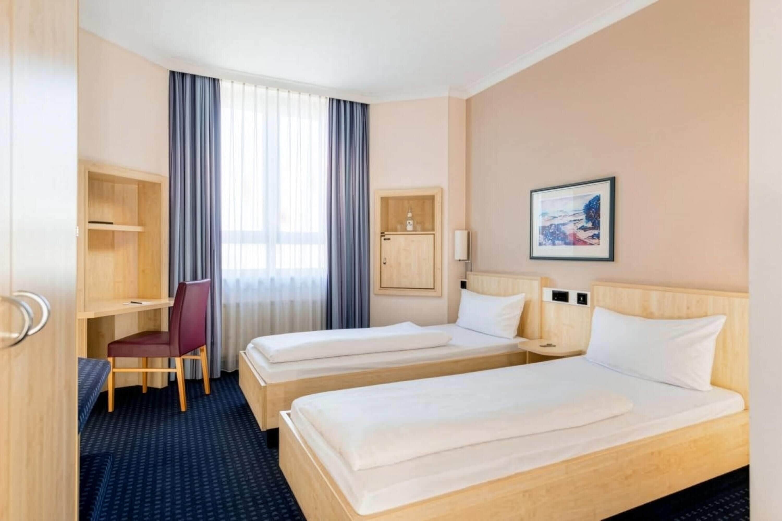 Quarto de hotel com duas camas, uma escrivaninha, uma cadeira e uma janela com cortinas.
