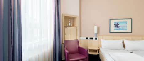 Quarto de hotel com cama, uma mesinha, uma cadeira, um criado-mudo e um quadro na parede.