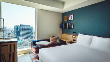 Quarto de hotel com uma cama grande, área de estar com vista para a cidade, uma pequena mesa e uma estante.