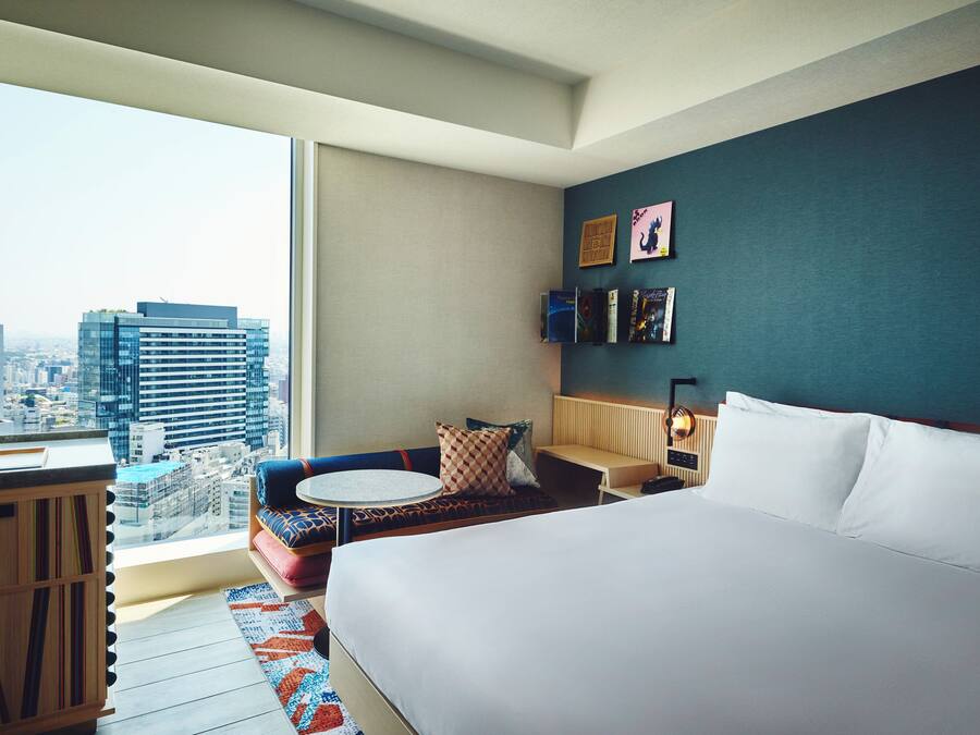 Quarto de hotel com uma cama grande, área de estar com vista para a cidade, uma pequena mesa e uma estante.