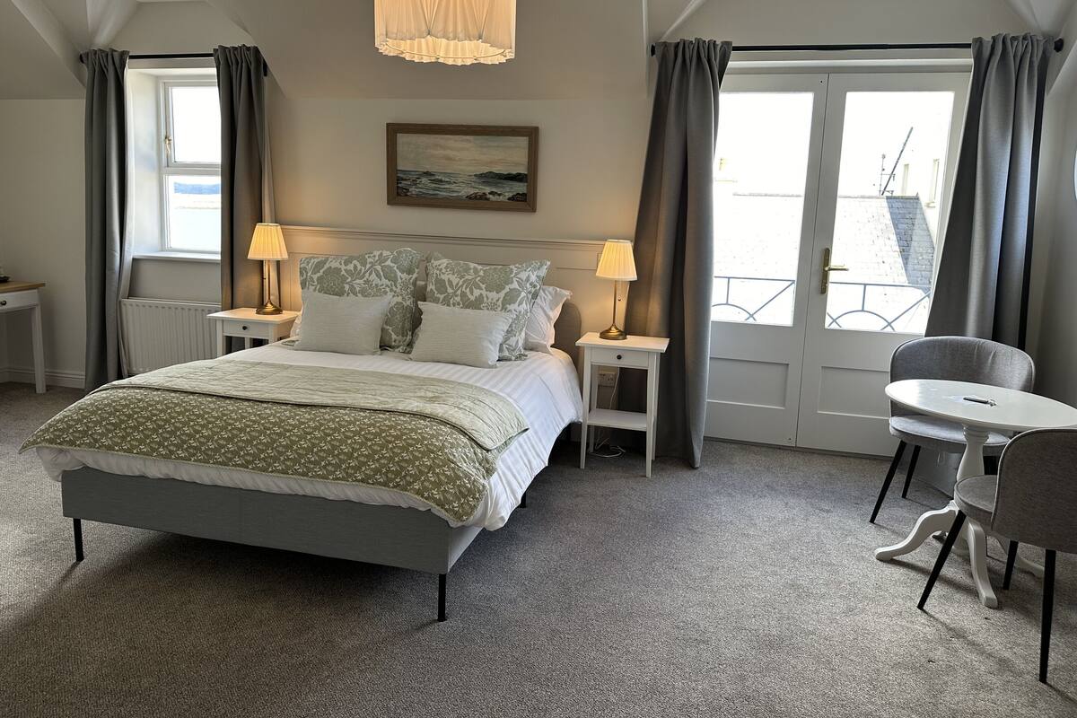Um quarto com uma cama, duas mesas de cabeceira com abajures, uma cadeira, uma janela com cortinas e um quadro na parede.