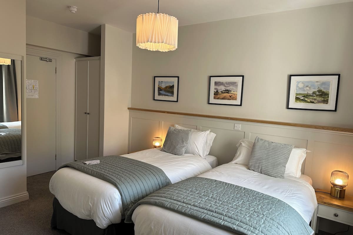 Quarto de hotel com duas camas, uma cabeceira de madeira, um lustre e três quadros na parede.