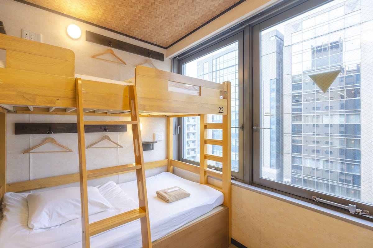 Quarto com beliches de madeira, janela com vista para edifícios e luminária pendente.