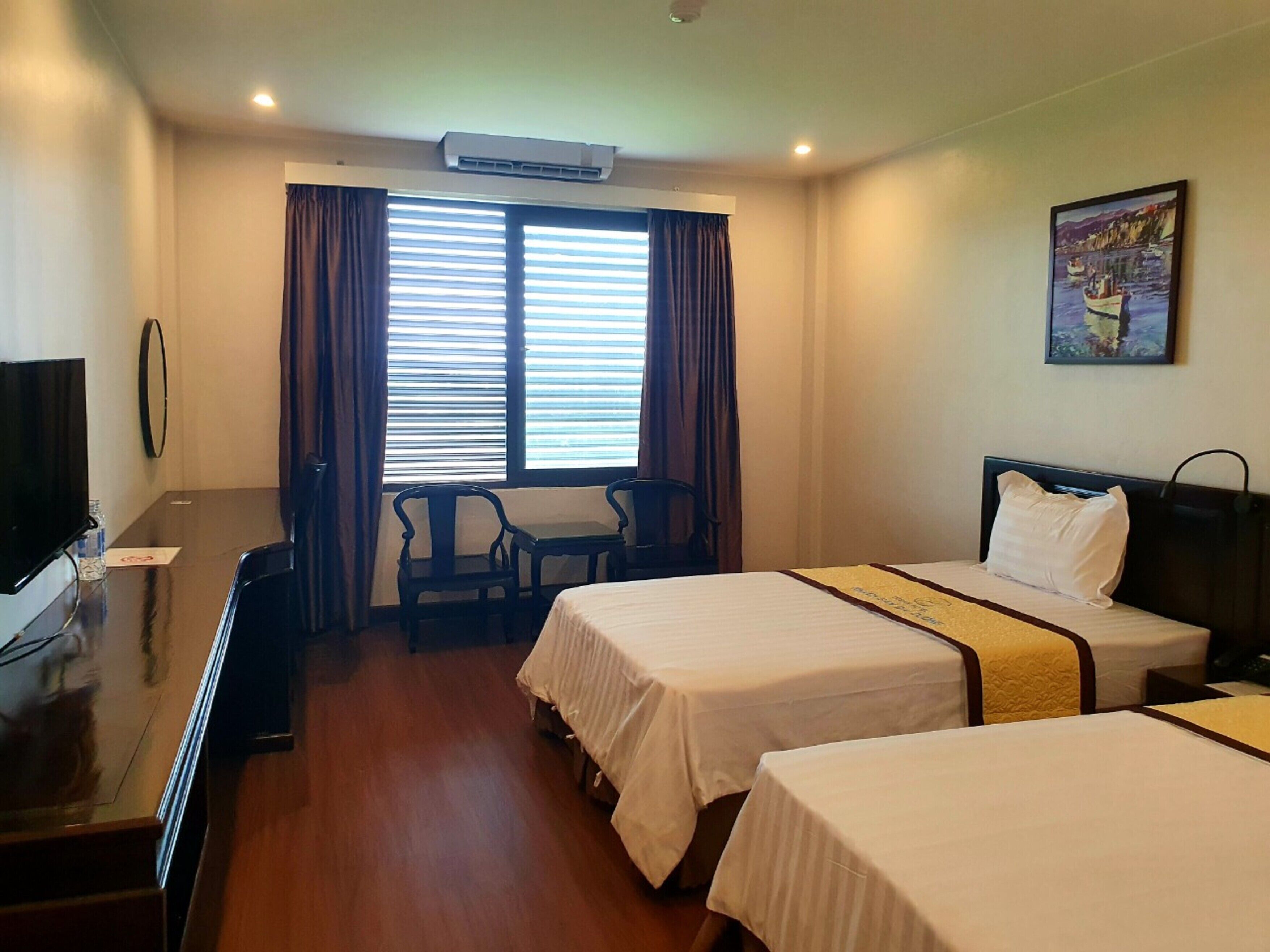 Quarto de hotel com duas camas, uma escrivaninha com televisão, uma janela com cortinas e um quadro na parede.