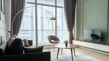 Uma sala de estar moderna com um sofá, poltrona e uma mesinha com uma planta. Uma televisão está instalada na parede, e há uma janela grande com cortinas.