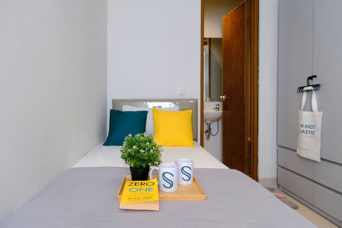 Cama bem arrumada com uma planta em vaso, um tabuleiro com duas chávenas e um livro intitulado 