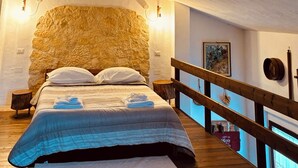 Quarto com cabeceira de pedra, piso de madeira, cama com roupa de cama azul e corrimão de madeira.