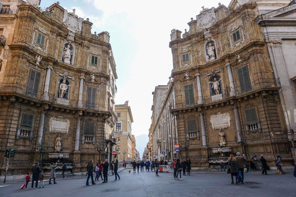 Rua com edifícios ornamentados e arquitetura clássica, incluindo estátuas.
