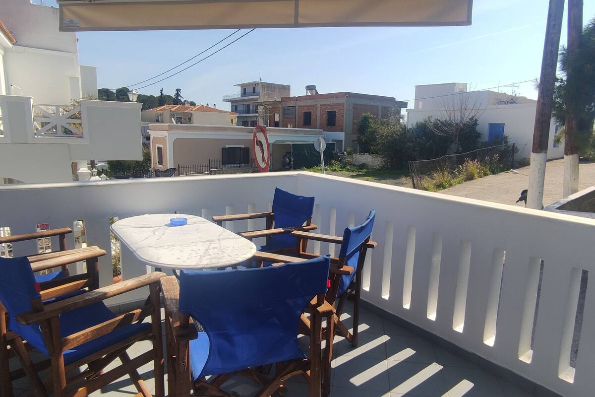 Uma varanda com mesa e cadeiras, grade branca e vista para edifícios e árvores.