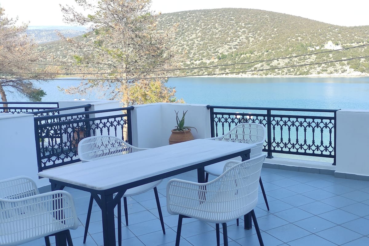 Varanda com mesa e cadeiras, um vaso de planta e vista para um lago e colinas.
