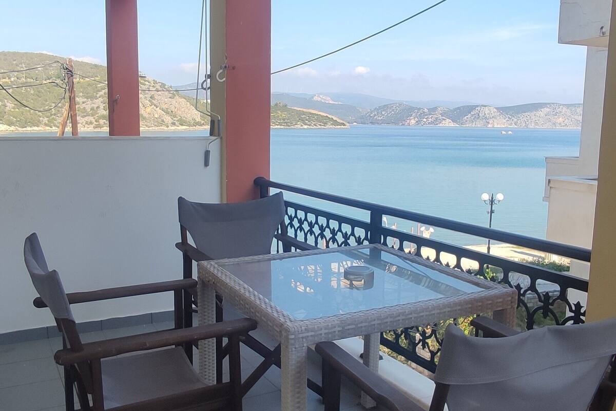 Uma varanda com mesa de vidro e quatro cadeiras de madeira, com vista para a paisagem costeira.