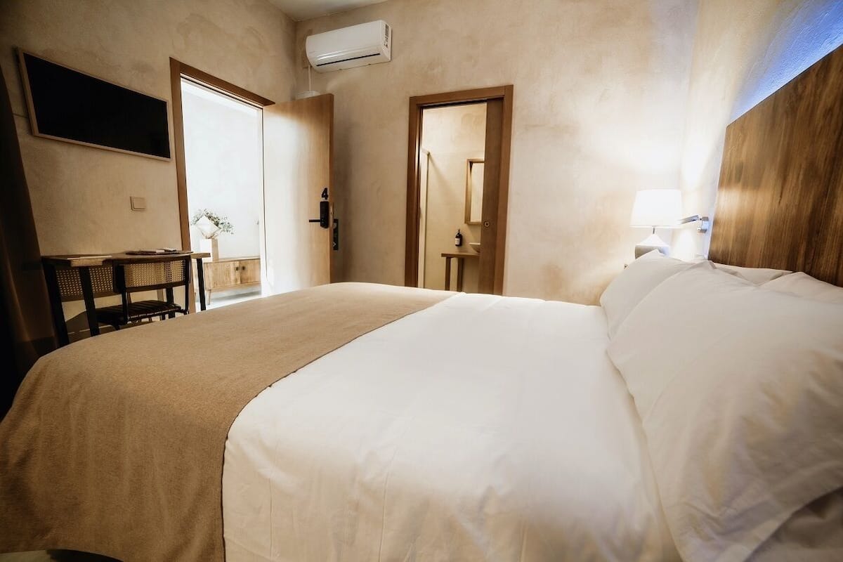 Quarto de hotel com cama, escrivaninha, cadeira, espelho e porta para o banheiro.