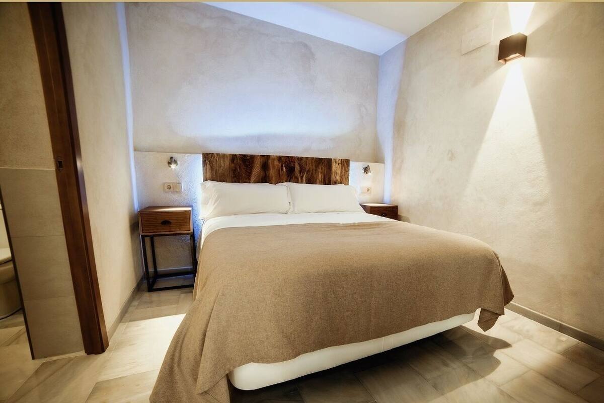 Quarto com cama, mesas de cabeceira, cabeceira de madeira e luminária fixada na parede.