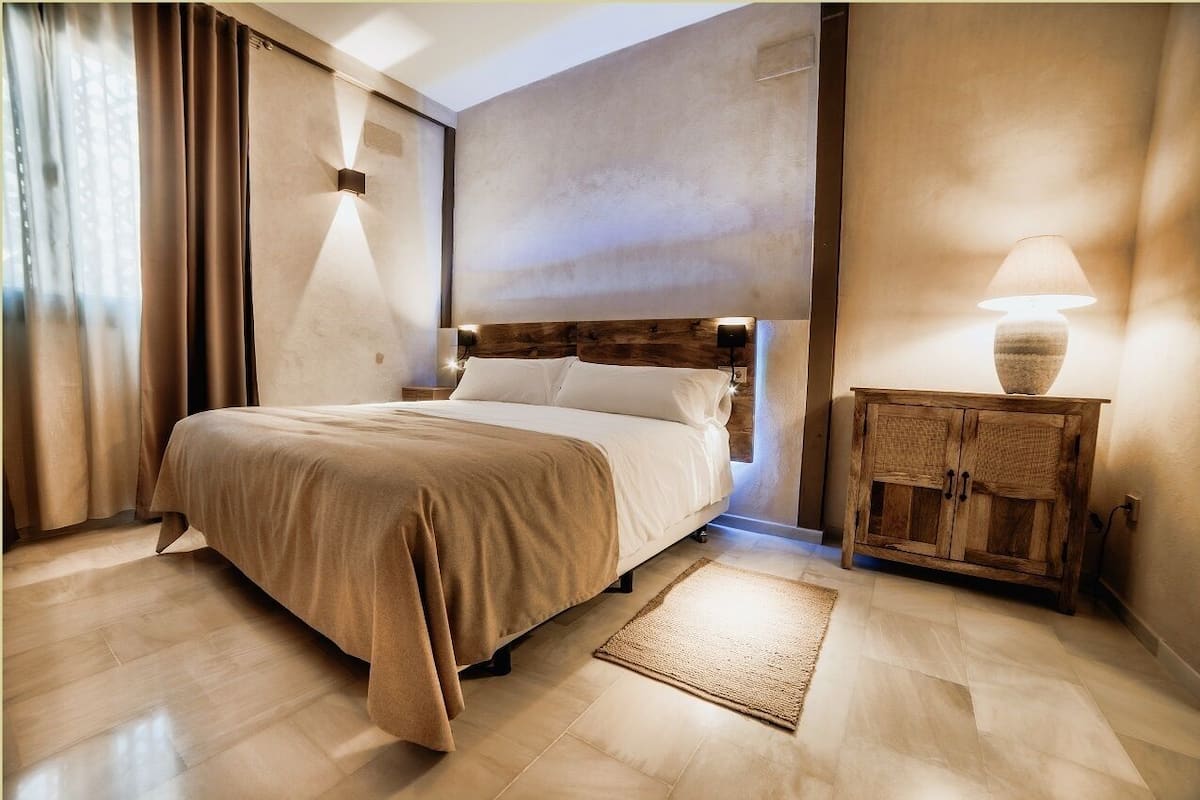 Quarto bem arrumado com uma cama grande, uma mesa de cabeceira de madeira com abajur e um pequeno armário de madeira.