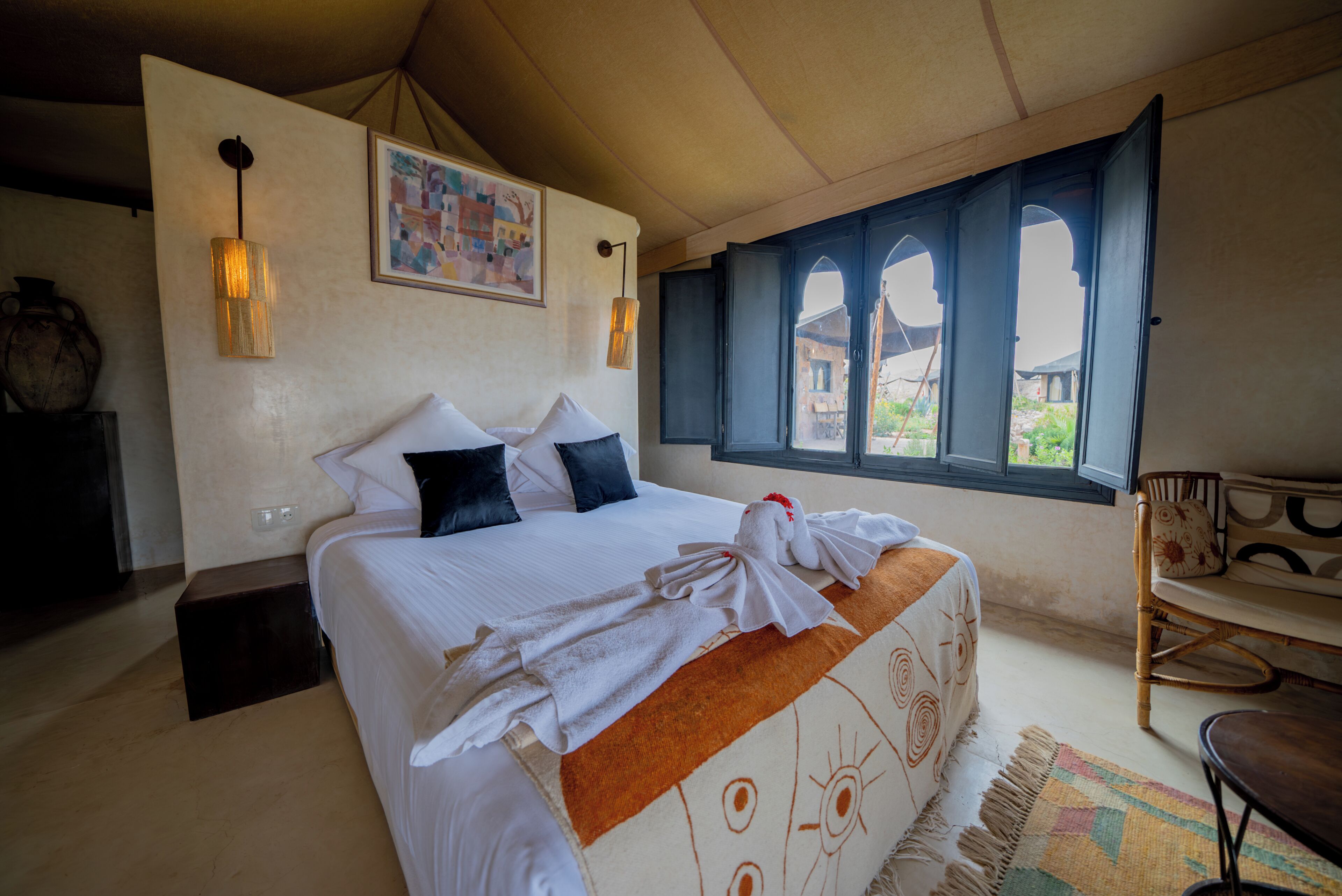 Uma cama bem arrumada com travesseiros e cobertor, um criado-mudo de madeira, uma luminária fixada na parede e uma janela com vista para prédios.