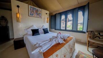 Uma cama bem arrumada com travesseiros e cobertor, um criado-mudo de madeira, uma luminária fixada na parede e uma janela com vista para prédios.