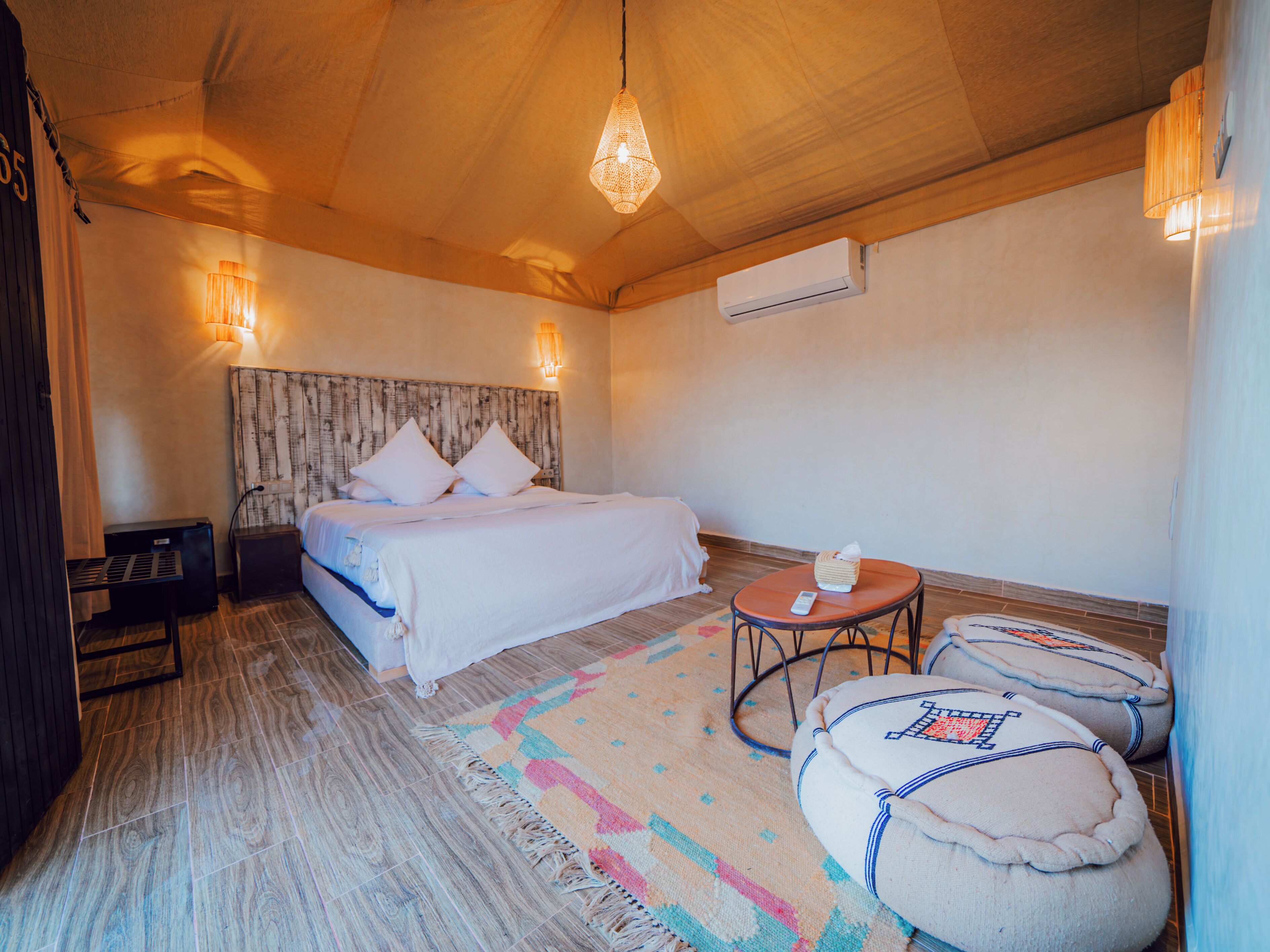 Quarto de hotel com cama, mesa de cabeceira, luminária, aparelho de ar condicionado e uma pequena mesa redonda com um controle remoto.