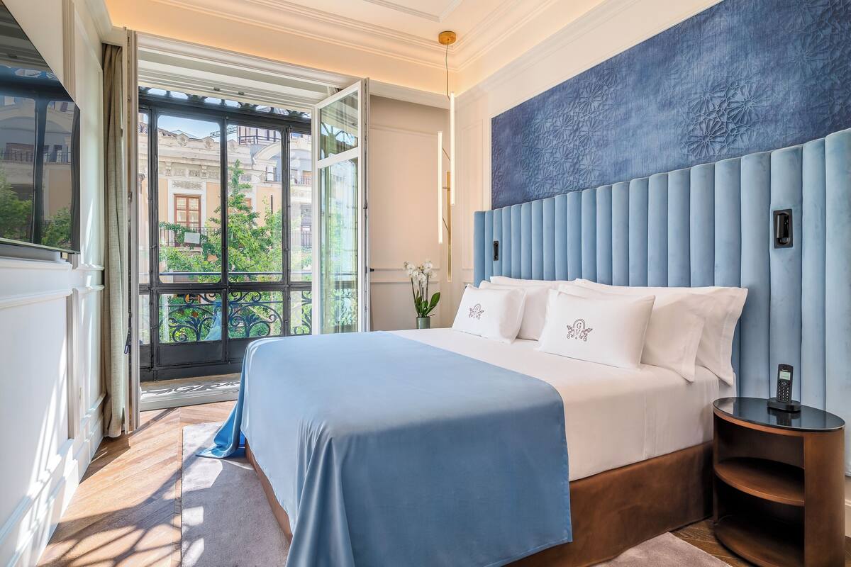 Quarto com uma cama grande, cabeceira azul, mesa de cabeceira e vista para a varanda.