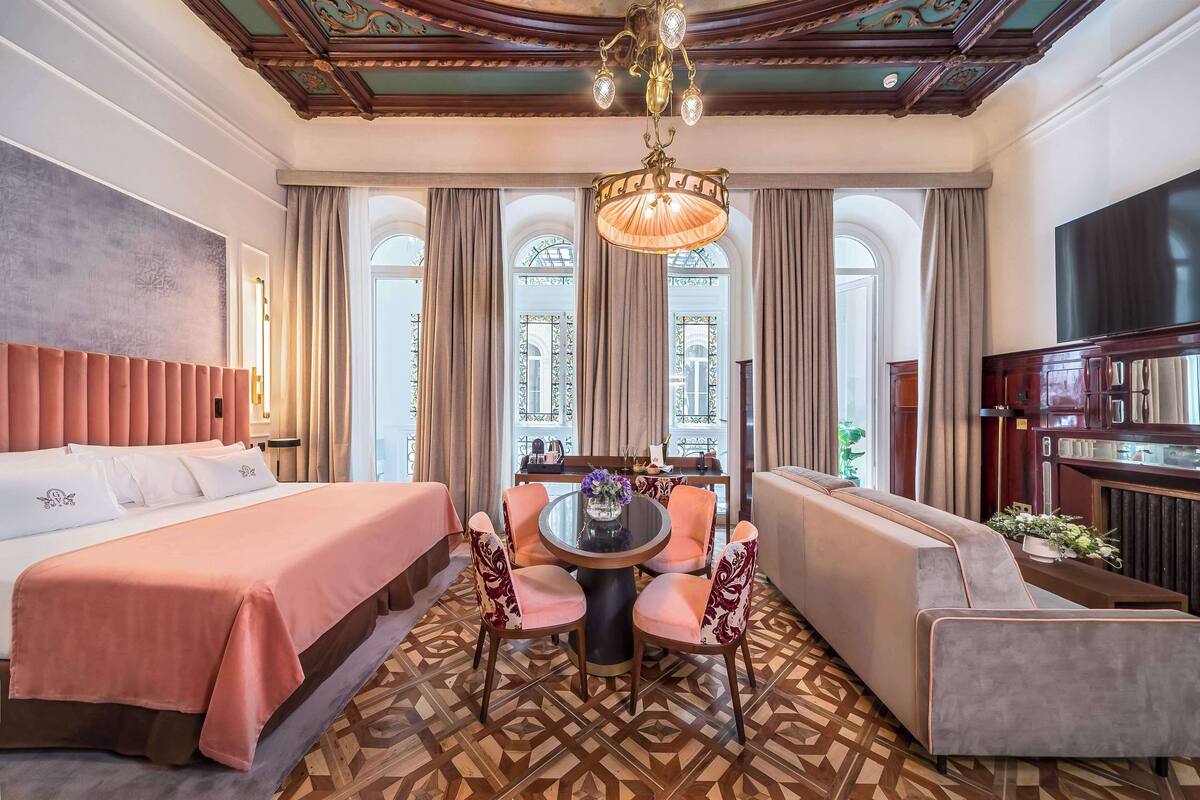 Quarto de hotel com uma cama grande, um sofá, uma mesa de jantar com cadeiras e um candelabro.