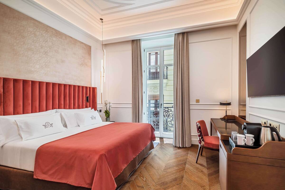 Quarto de hotel com uma cama grande, uma mesa com cadeira, televisão e varanda com guarda-corpo.