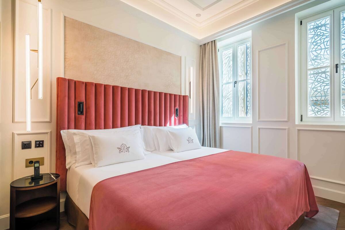 Quarto de hotel com cama grande, cabeceira vermelha, roupa de cama branca e telefone ao lado da cama.
