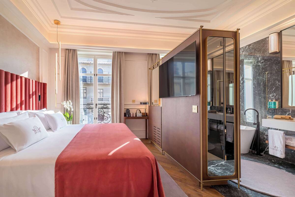 Quarto de hotel com uma cama grande, banheiro com bancada de mármore e vista para a cidade pela janela.