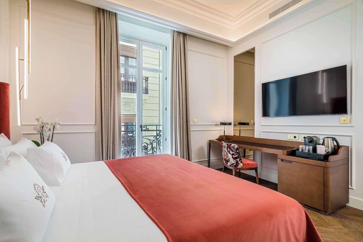 Quarto de hotel com cama, escrivaninha, televisão e varanda com cortinas.