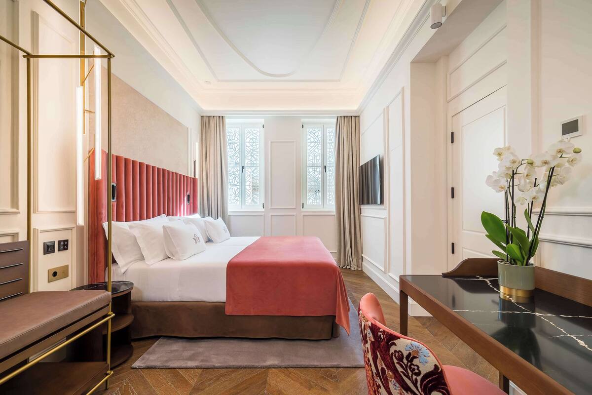 Quarto de hotel com cama, mesa de cabeceira, cadeira, televisão e janela com cortinas.