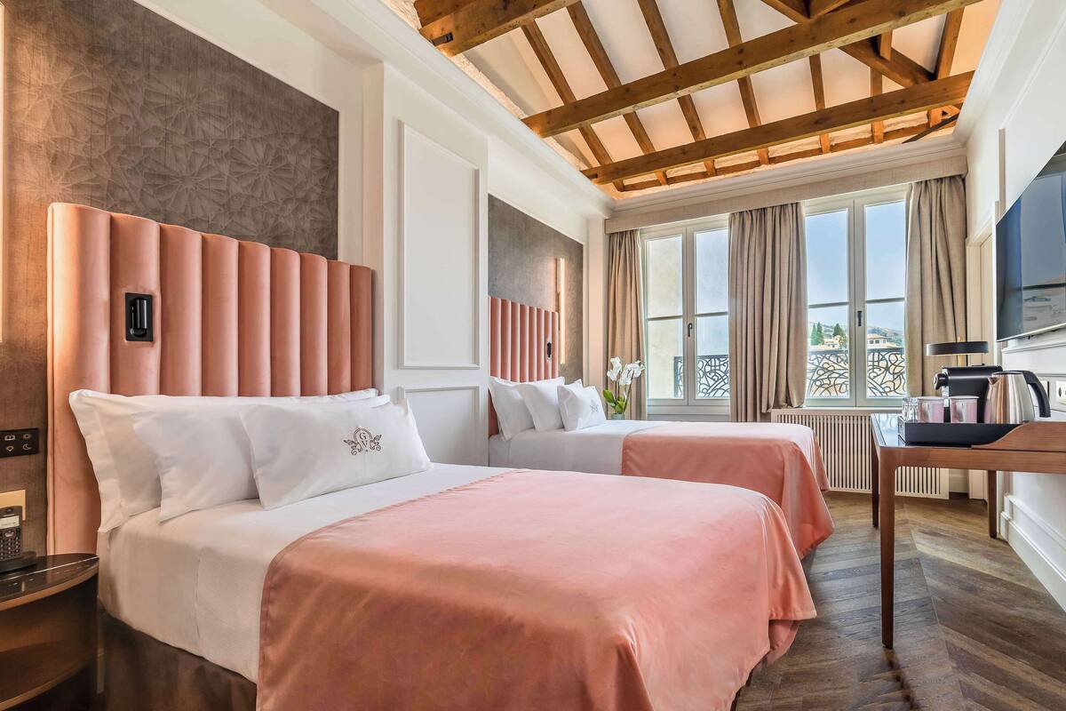 Quarto de hotel com duas camas, teto de madeira, uma janela grande com cortinas e uma televisão em uma mesa de madeira.