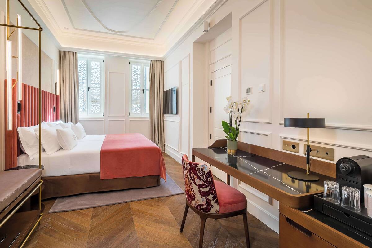 Quarto de hotel com cama, escrivaninha com cafeteira, cadeira e janela com cortinas.