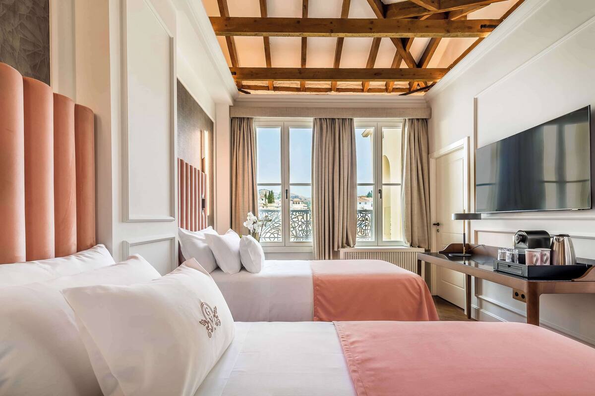 Quarto de hotel com duas camas, uma escrivaninha com cafeteira, TV e varanda com vista.