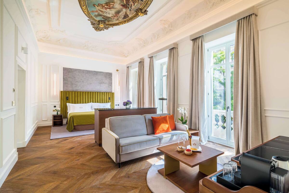 Quarto de hotel com sofá, mesa de centro, cama com cabeceira verde e vista para o exterior por grandes janelas.