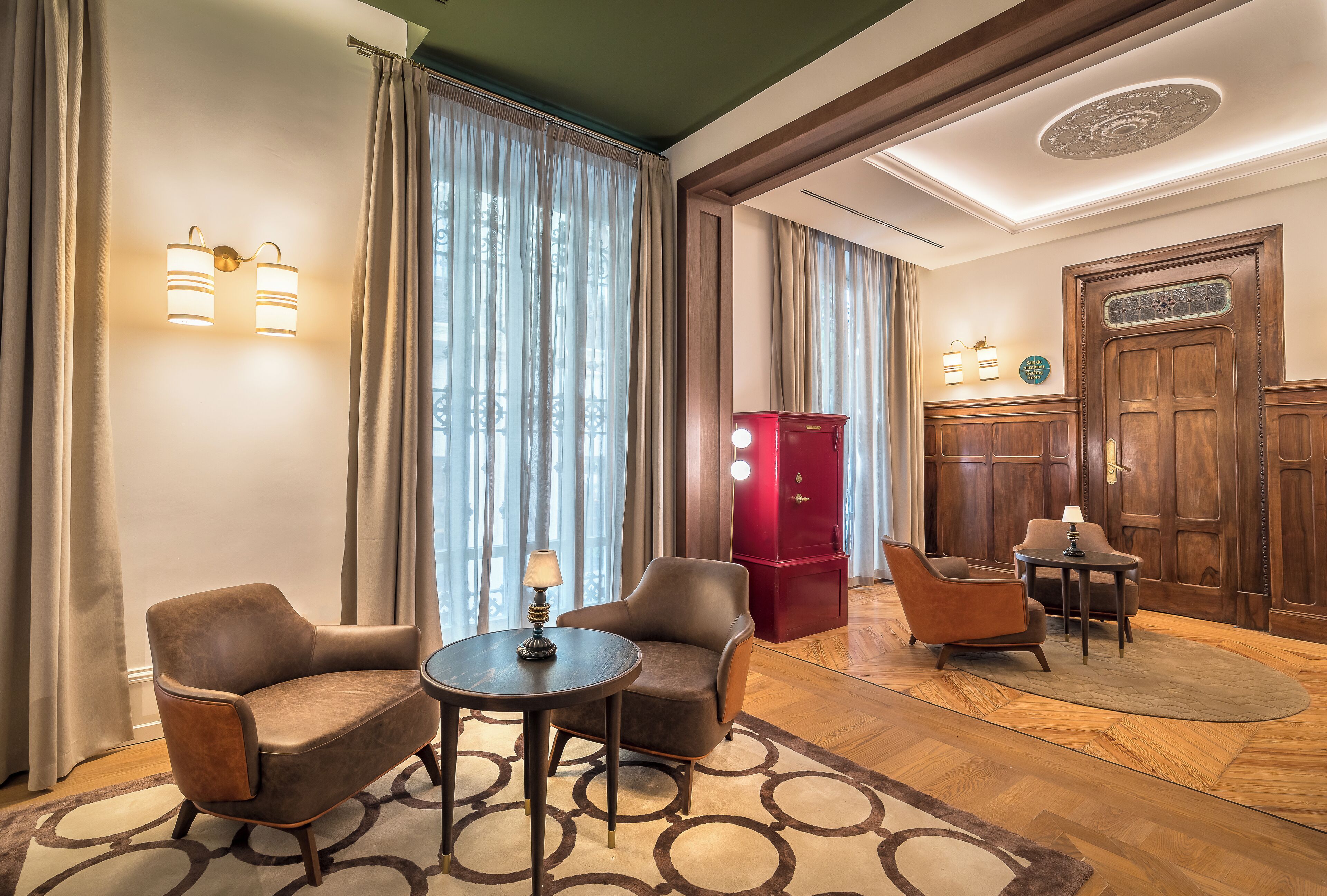 Quarto de hotel com área de estar, incluindo duas poltronas e uma pequena mesa, um armário vermelho e uma porta de madeira com painel de vidro decorativo.
