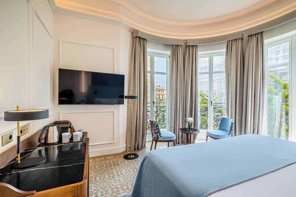 Quarto de hotel com uma cama, uma escrivaninha com abajur, uma televisão e vista da cidade através de grandes janelas.