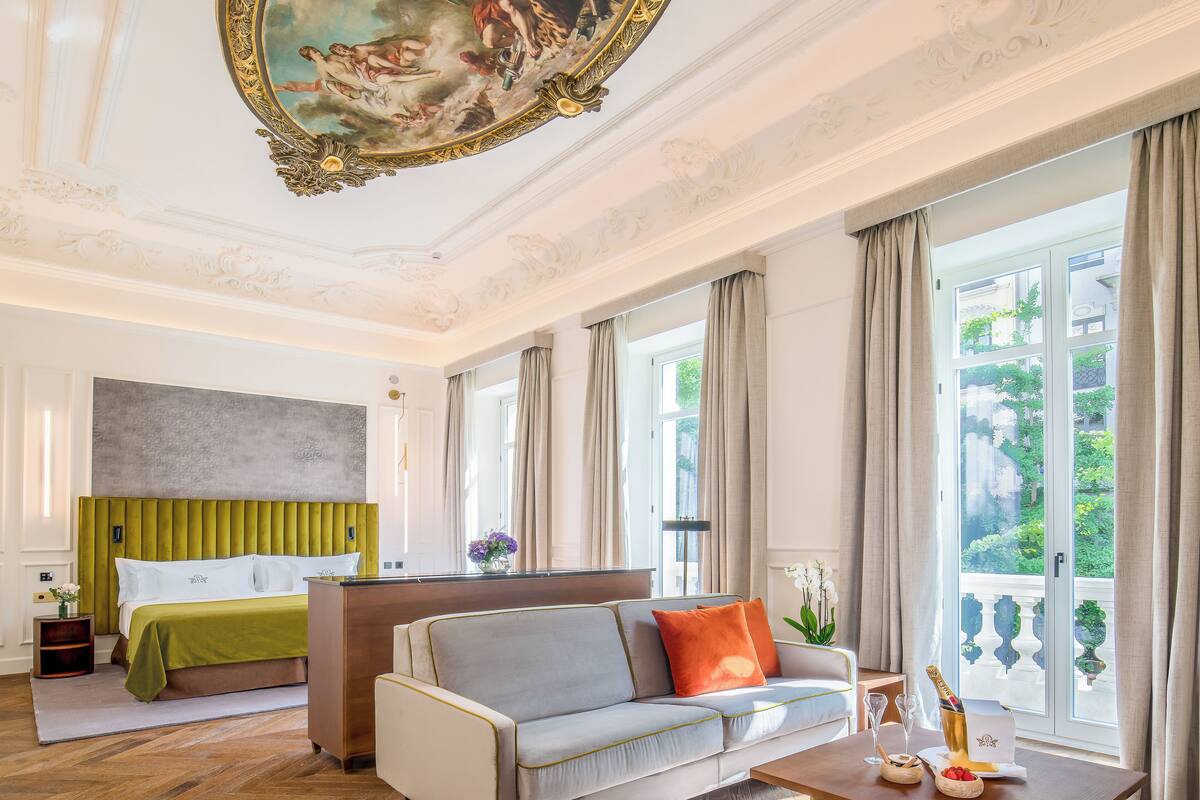 Quarto espaçoso com uma cama grande, um sofá e uma mesa de jantar. Há vista para o verde pelas janelas.