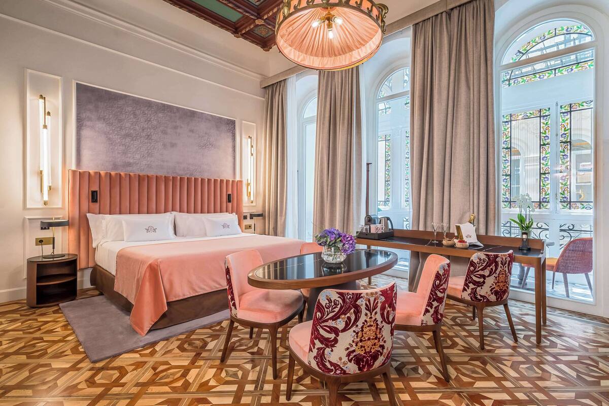 Quarto de hotel com uma cama grande, uma mesa de jantar redonda e vista para o exterior através de amplas janelas.