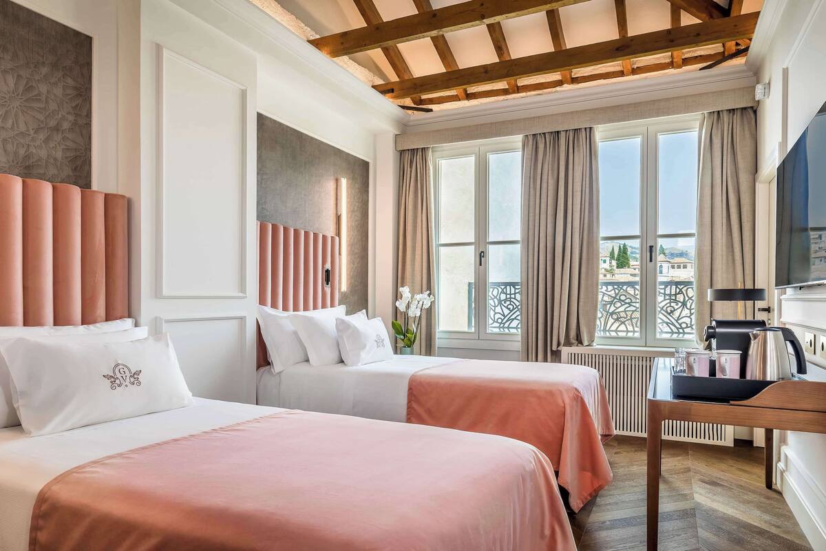 Quarto de hotel com duas camas, teto de madeira, uma janela grande com cortinas, um radiador e uma pequena mesa com uma chaleira.