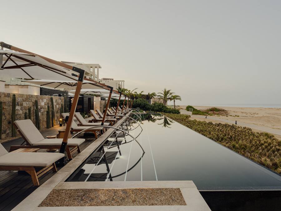 Uma piscina infinita com espreguiçadeiras, vista para a praia e paisagem desértica.