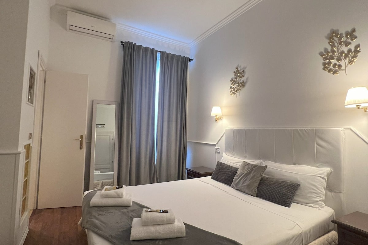 Quarto de hotel com uma cama grande, uma mesa de cabeceira, um espelho, uma janela com cortinas e um ar condicionado fixado na parede.