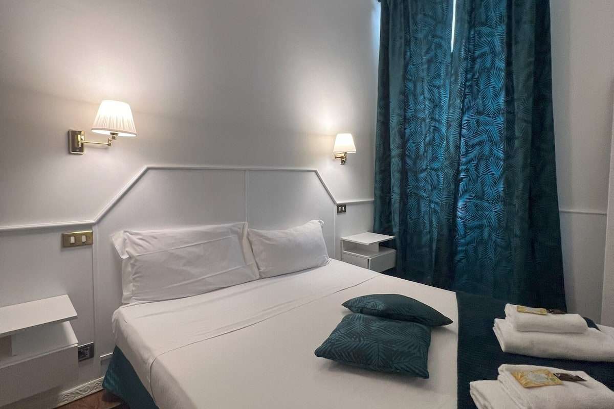 Cama bem arrumada com roupa de cama branca e azul-turquesa, duas luminárias de cabeceira, mesa de cabeceira e janela com cortinas azul-turquesa.