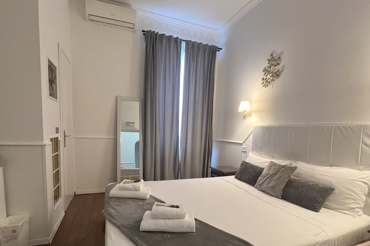 Quarto de hotel com uma cama grande, uma mesa de cabeceira, um espelho, uma janela com cortinas e um aparelho de ar condicionado.