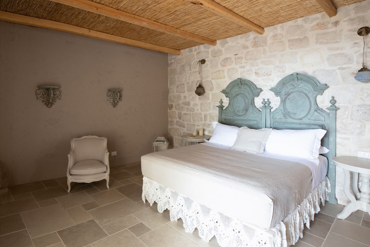 Quarto com parede de pedra, teto de madeira, cama com cabeceira branca, uma cadeira e uma mesa pequena.