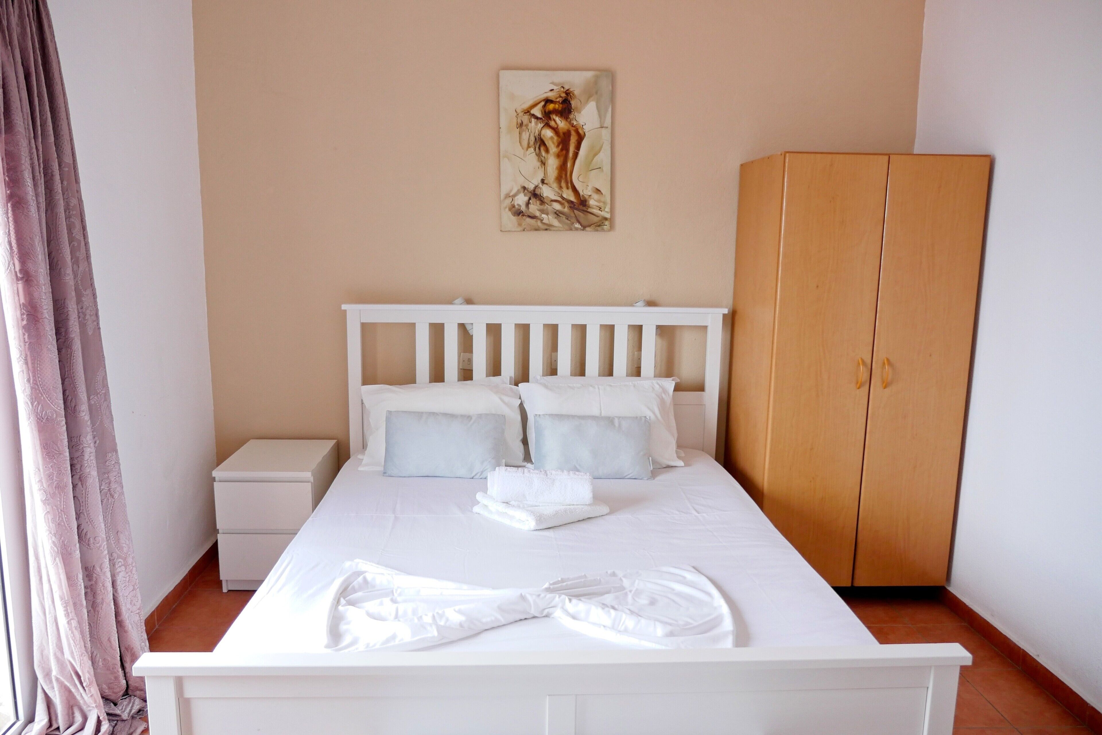 Quarto com cama branca, duas almofadas, mesa de cabeceira, guarda-roupa de madeira e um quadro na parede.