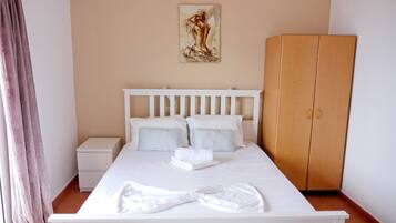 Quarto com cama branca, duas almofadas, mesa de cabeceira, guarda-roupa de madeira e um quadro na parede.