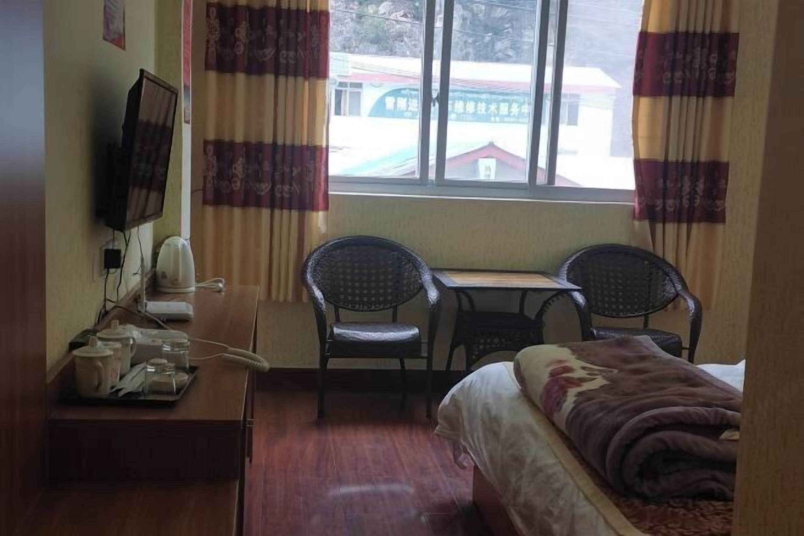 Quarto de hotel com uma cama, uma escrivaninha com telefone, duas cadeiras, uma televisão e uma janela com cortinas.