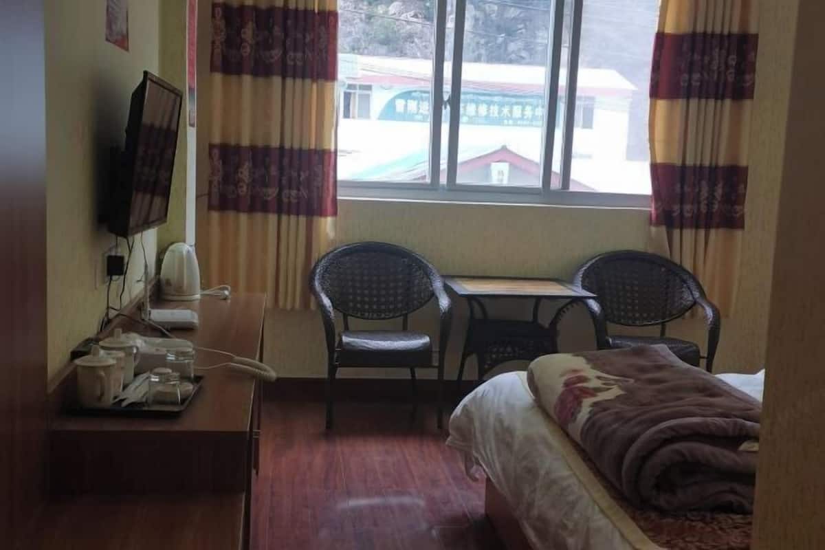 Quarto de hotel com uma cama, uma escrivaninha com telefone, duas cadeiras, uma televisão e uma janela com cortinas.