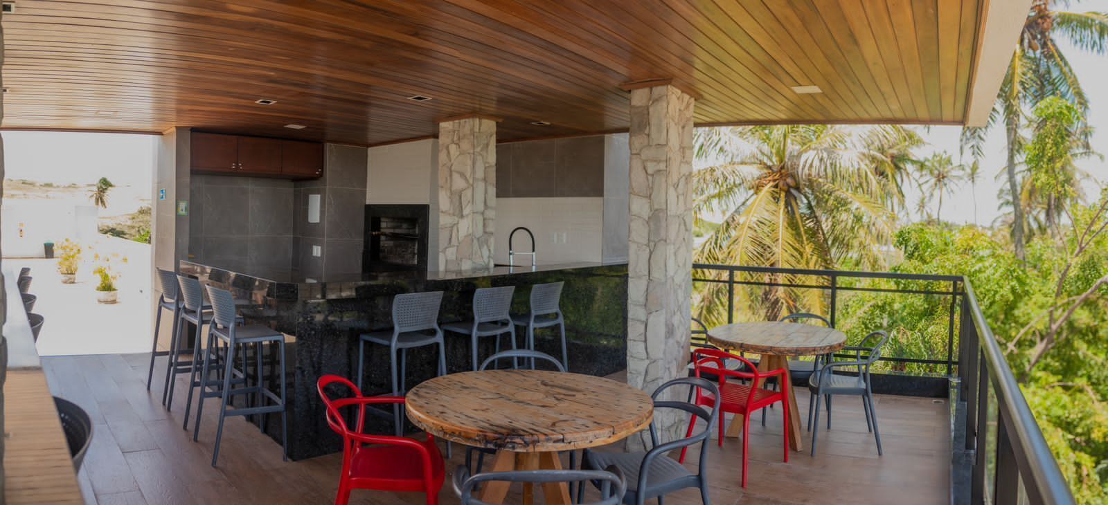 Uma área de cozinha externa com um pilar de pedra, churrasqueira embutida e um espaço bar com banquetas. Há mesas e cadeiras redondas ao redor do pátio.
