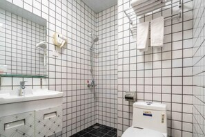 Uma casa de banho pequena com azulejos, uma pia branca, uma prateleira de vidro, um chuveiro e uma sanita.